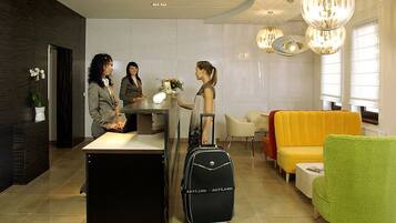 Uma área de recepção de hotel com balcão, sala de espera com sofá e cadeiras, e uma mala preta grande.