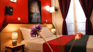 Quarto de hotel com parede vermelha, cama com roupa de cama branca, colcha vermelha e preta, mesa de cabeceira com abajur, quadro com a Torre Eiffel e janela com cortinas vermelhas.