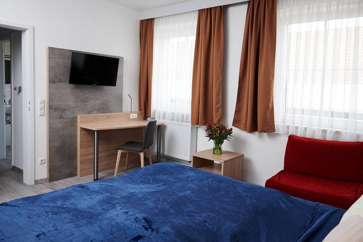 Quarto de hotel com cama, escrivaninha, cadeira, televisão fixada na parede e janela com cortinas.