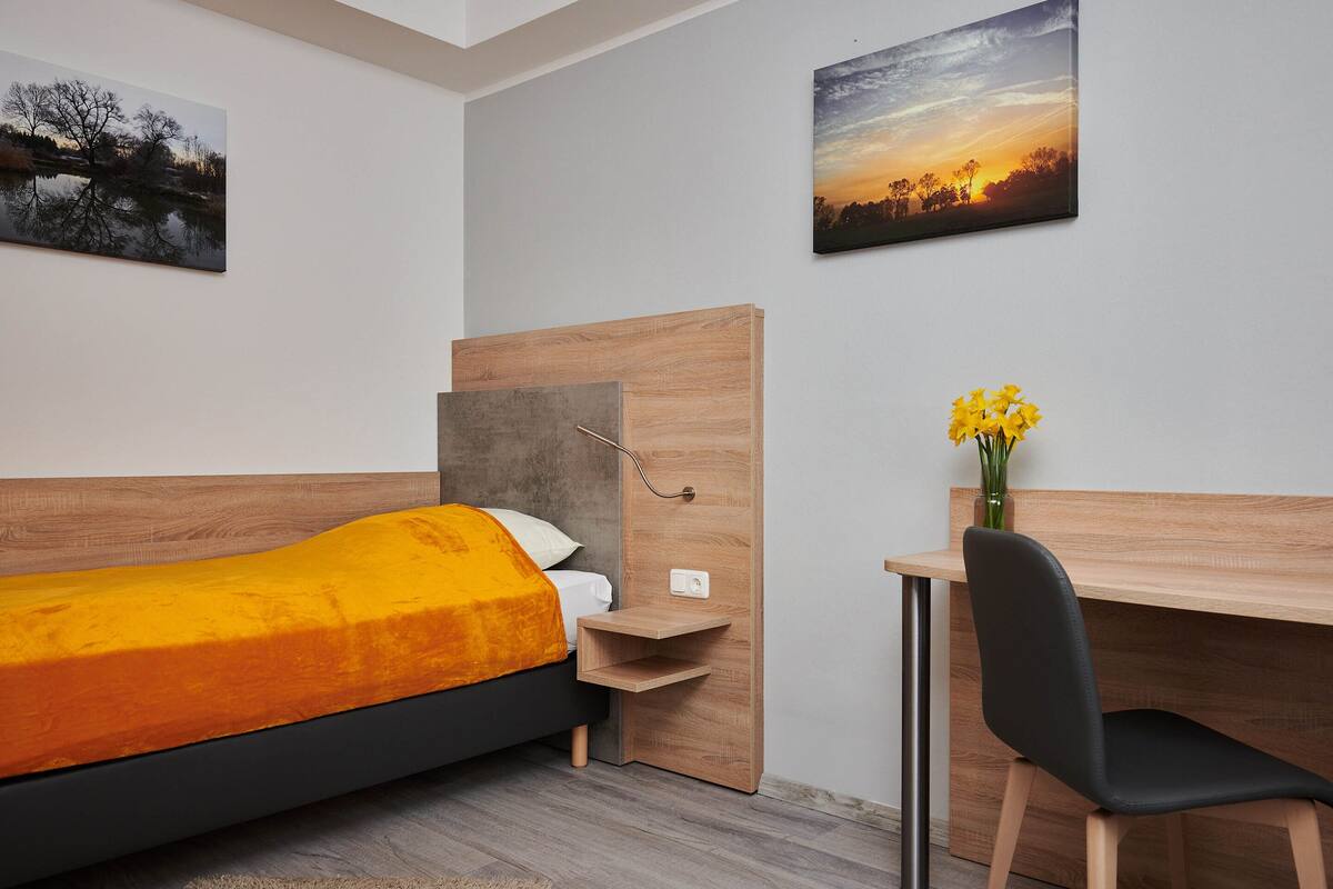 Quarto com cama, escrivaninha e cadeira. Há duas pinturas na parede.