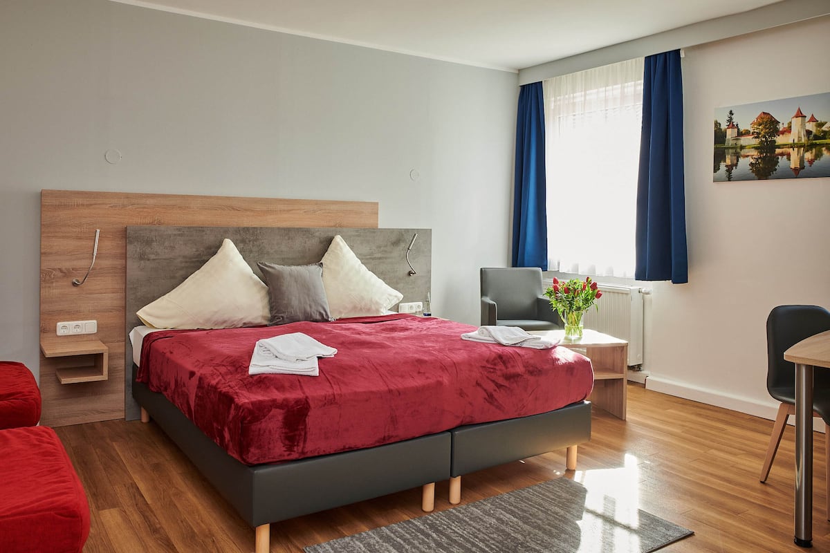 Quarto moderno com uma cama grande, uma mesinha de cabeceira, uma cadeira e vista para um castelo pela janela.