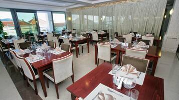 Uma sala de jantar com várias mesas preparadas para o serviço, grandes janelas e vista para o exterior.
