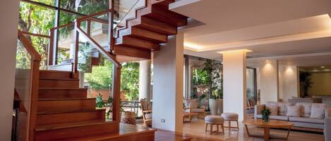 Interior moderno com escada de madeira, sala de estar com sofá e poltronas, e janelas amplas com vista para o verde.