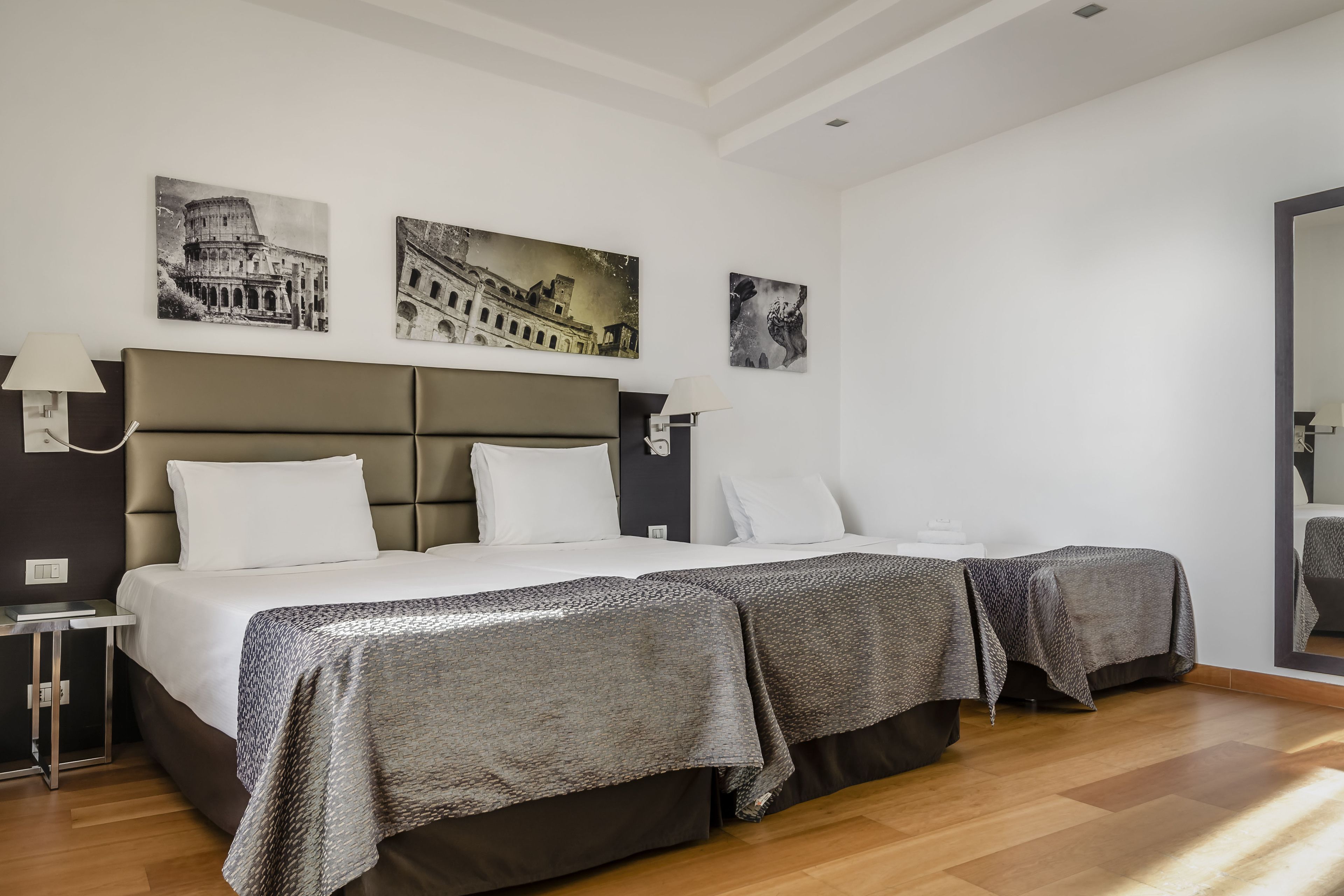Quarto de hotel com uma cama grande, duas mesas de cabeceira e um espelho. Há três quadros na parede acima da cama.