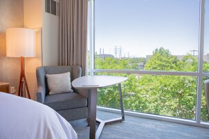 Quarto de hotel com cama, poltrona cinza com almofada, mesinha, luminária e uma grande janela com vista para áreas verdes e edifícios.