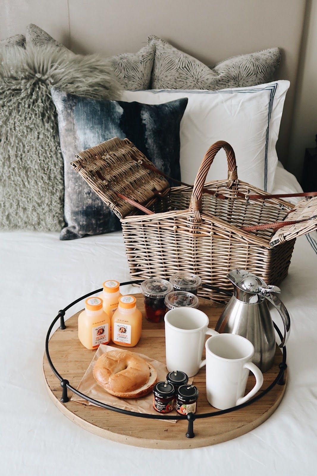 Bandeja de café da manhã com bagel, café, chá e acompanhamentos sobre a cama, ao fundo uma cesta de vime e almofadas.