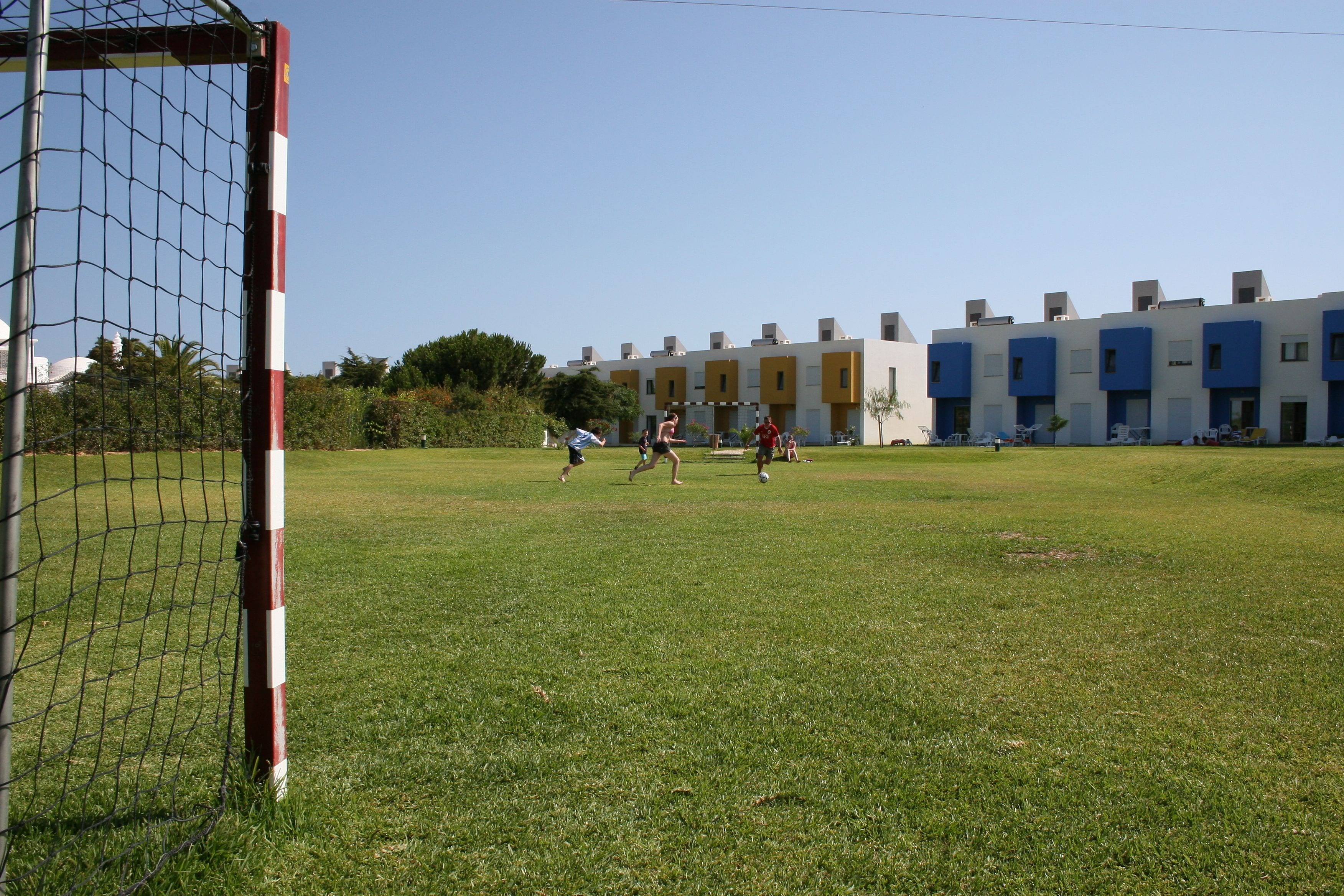 Um campo de futebol com um poste de gol, um edifício com várias varandas ao fundo e um céu claro.