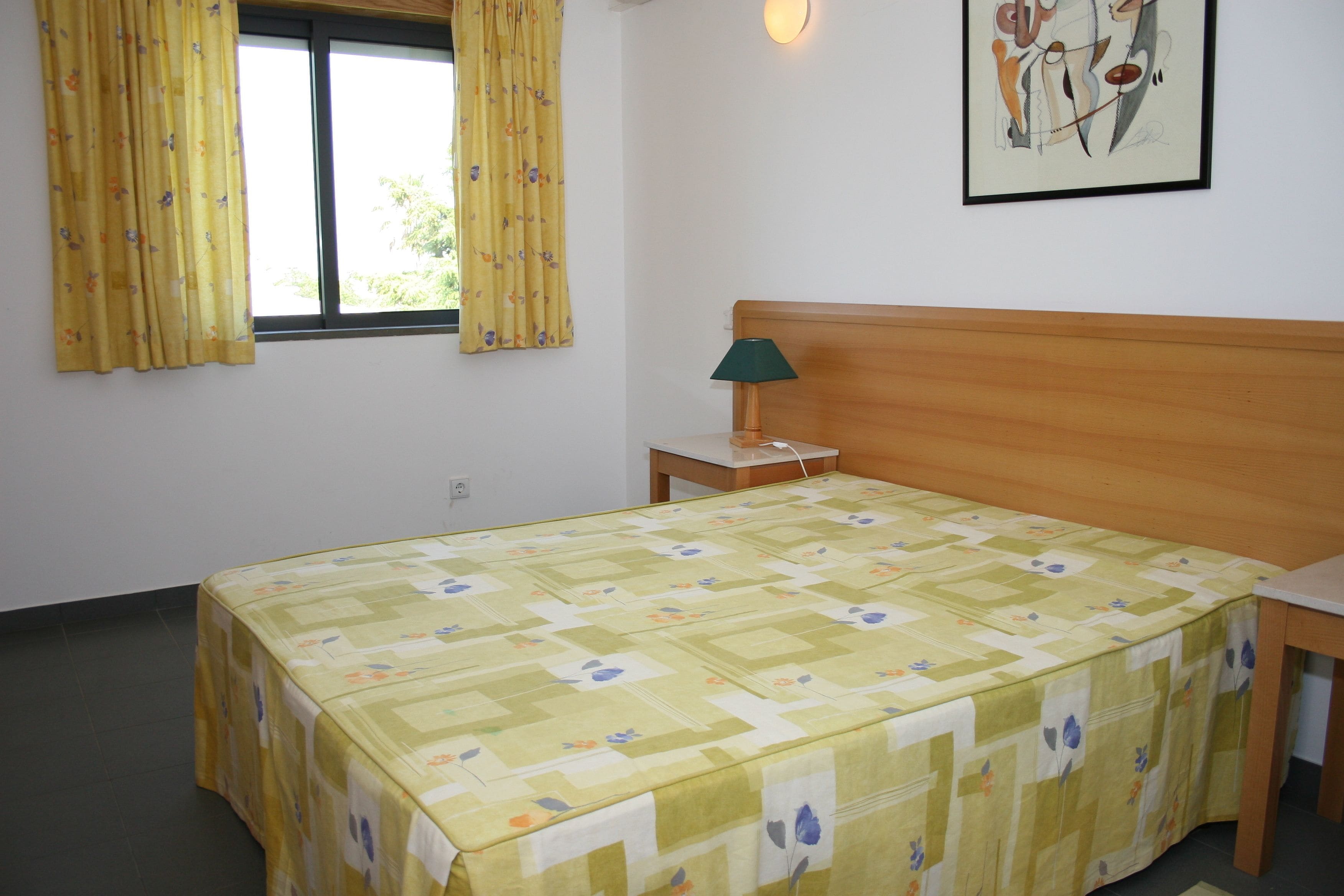 Quarto com cama, mesa de cabeceira, luminária, janela com cortinas e um quadro na parede.