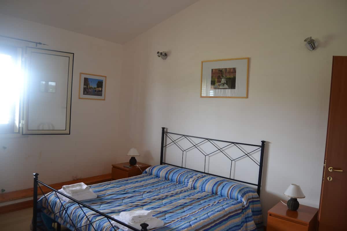 Um quarto com cama, duas mesas de cabeceira, um criado-mudo, uma porta e duas molduras com quadros na parede.