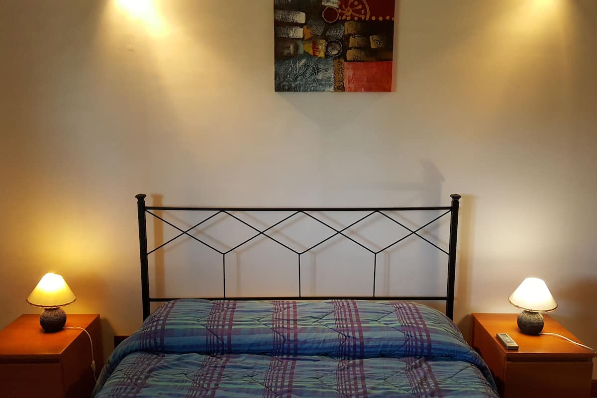Uma cama com cabeceira geométrica, duas luminárias de cabeceira, uma colcha estampada e um quadro na parede.
