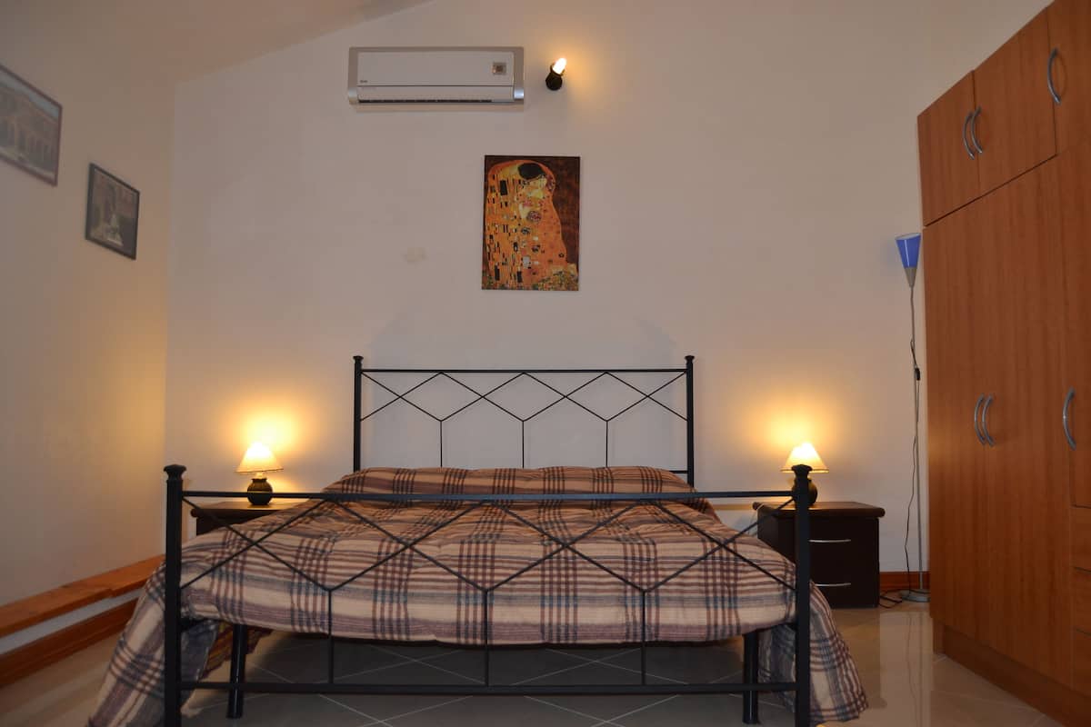 Quarto com cama, duas luminárias de apoio, ar-condicionado fixado na parede, um quadro e guarda-roupa de madeira.