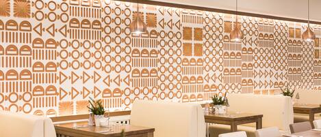 Interior de restaurante moderno com decoração de parede estampada, mesas de madeira e assentos acolchoados brancos.