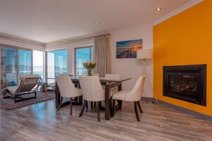 Uma sala de estar moderna com área de jantar, janelas amplas, lareira e vista da cidade.