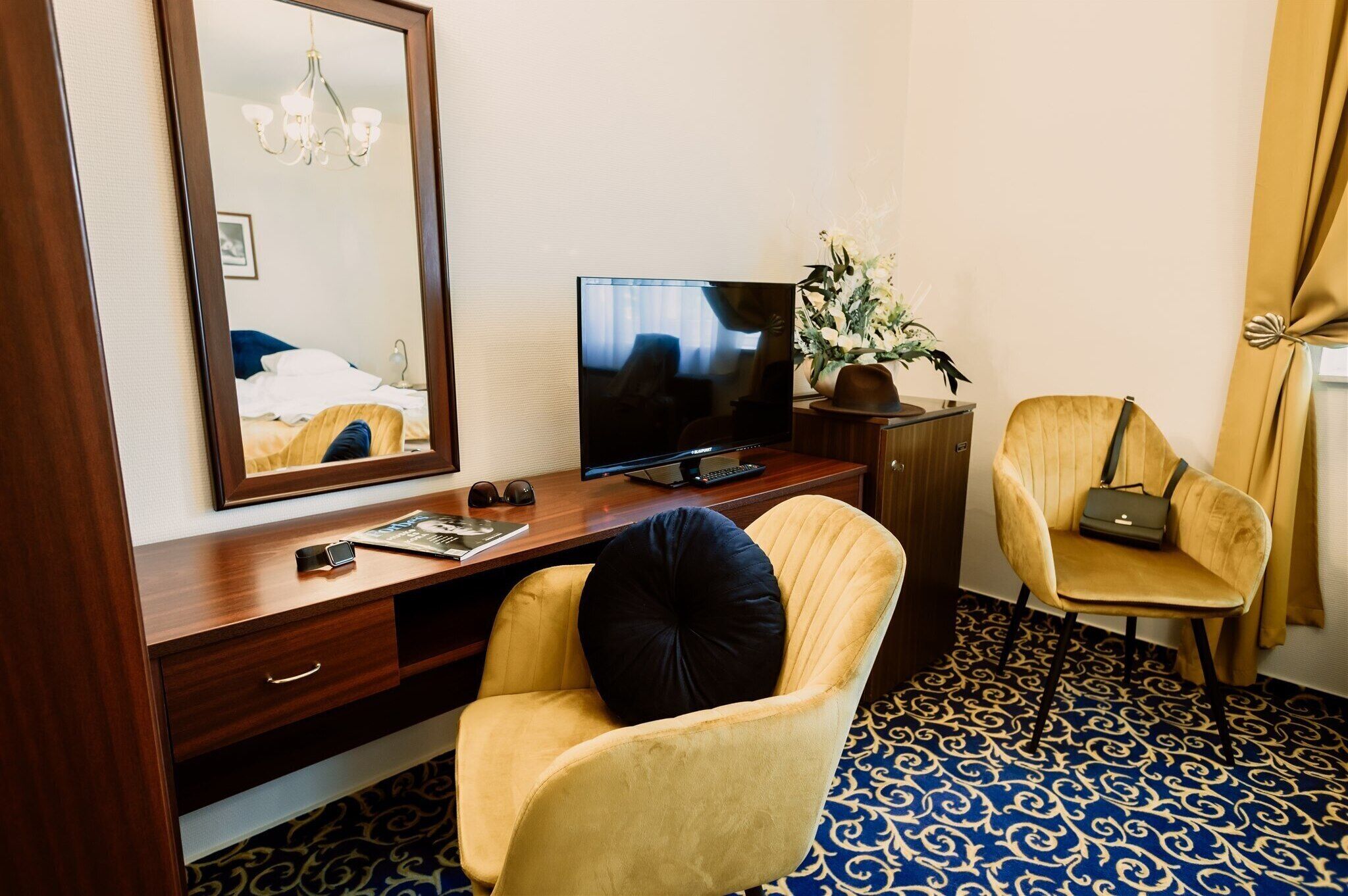 Quarto de hotel com escrivaninha de madeira, televisão, espelho, cadeira, mesa pequena com um vaso e tapete estampado.