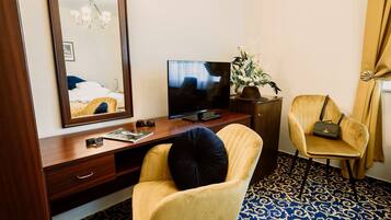 Quarto de hotel com escrivaninha de madeira, televisão, espelho, cadeira, mesa pequena com um vaso e tapete estampado.