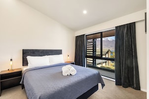 Quarto de hotel com cama, mesa de cabeceira, abajur, janela com cortinas e vista para as montanhas.