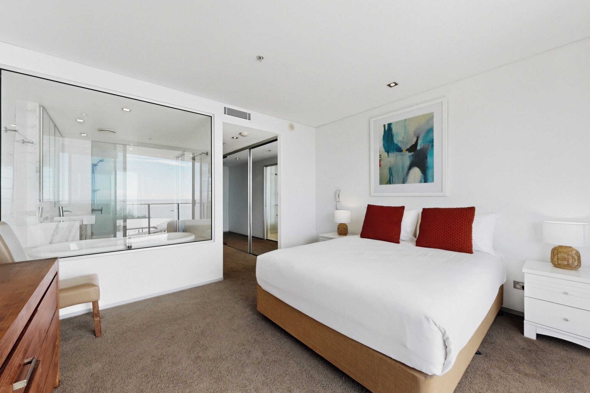 Quarto de hotel moderno com uma cama grande, uma escrivaninha, uma cadeira, banheiro com chuveiro de vidro e um quadro na parede.