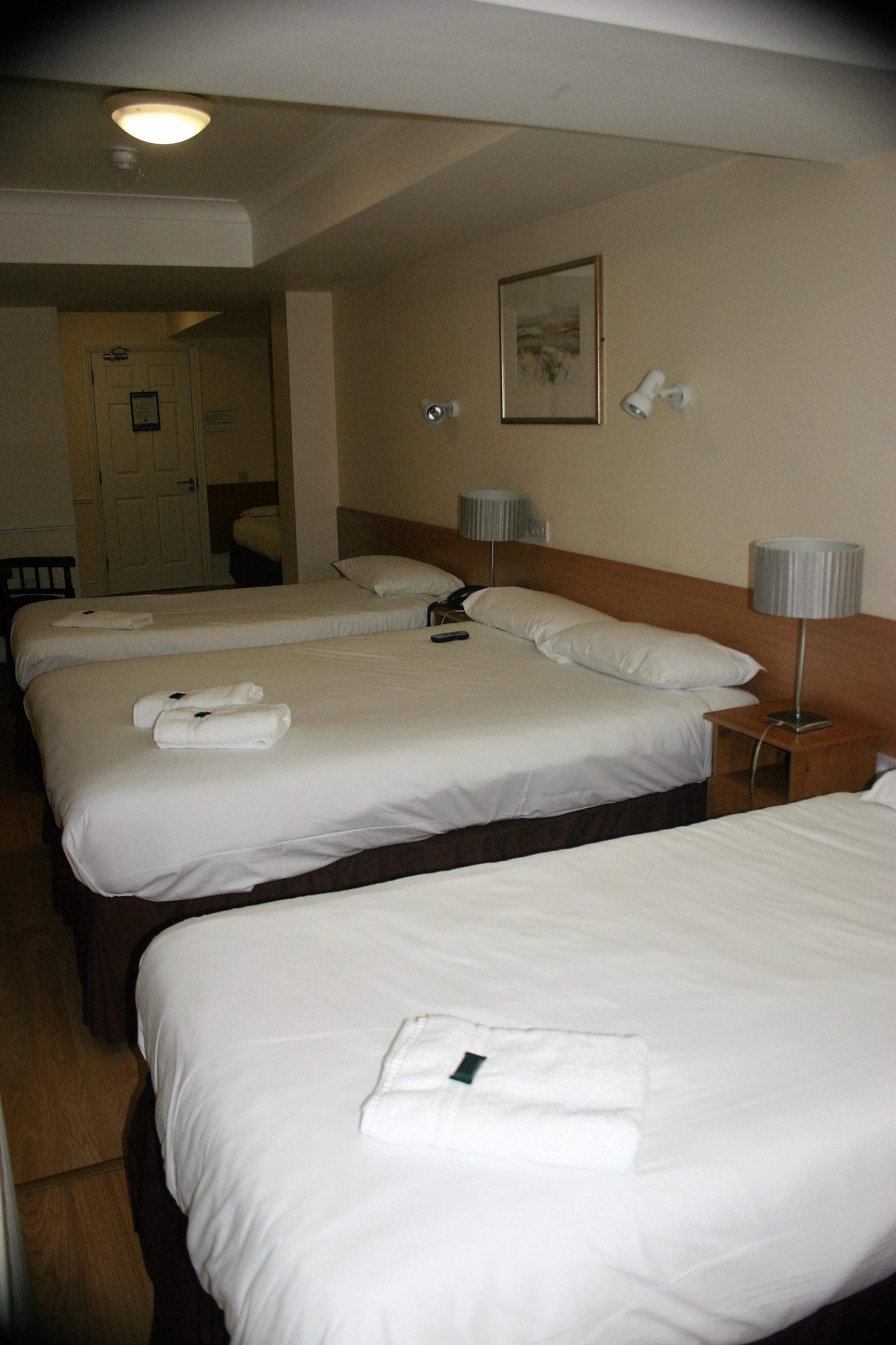 Quarto de hotel com três camas de solteiro, cada uma com uma toalha dobrada, uma mesa de cabeceira com abajur e um quadro na parede.