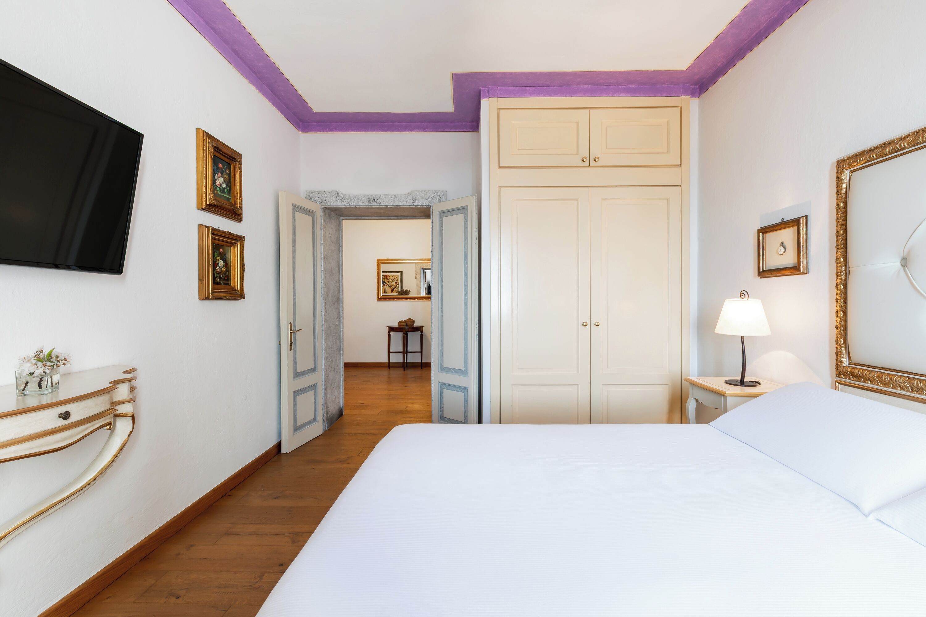 Quarto de hotel com uma cama, televisão, escrivaninha com candeeiro e guarda-roupa.