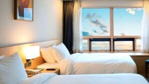 Quarto de hotel com duas camas, uma mesa de cabeceira com abajur, uma janela com vista para o céu e um quadro na parede.