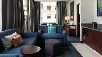 Quarto de hotel moderno com um sofá azul, um pequeno banquinho, uma mesa de centro redonda e vista para o horizonte urbano pelas grandes janelas.