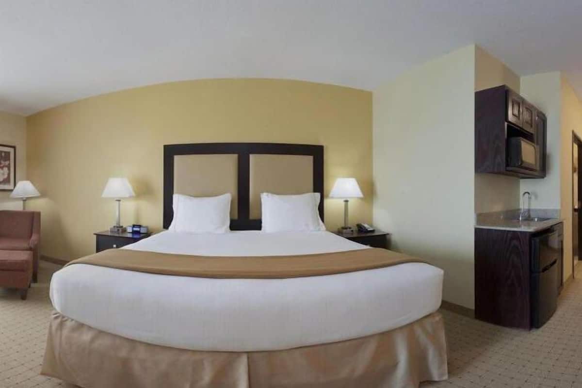 Quarto de hotel com uma cama grande, duas mesas de cabeceira com luminárias, uma cadeira, uma escrivaninha com pia e uma televisão.