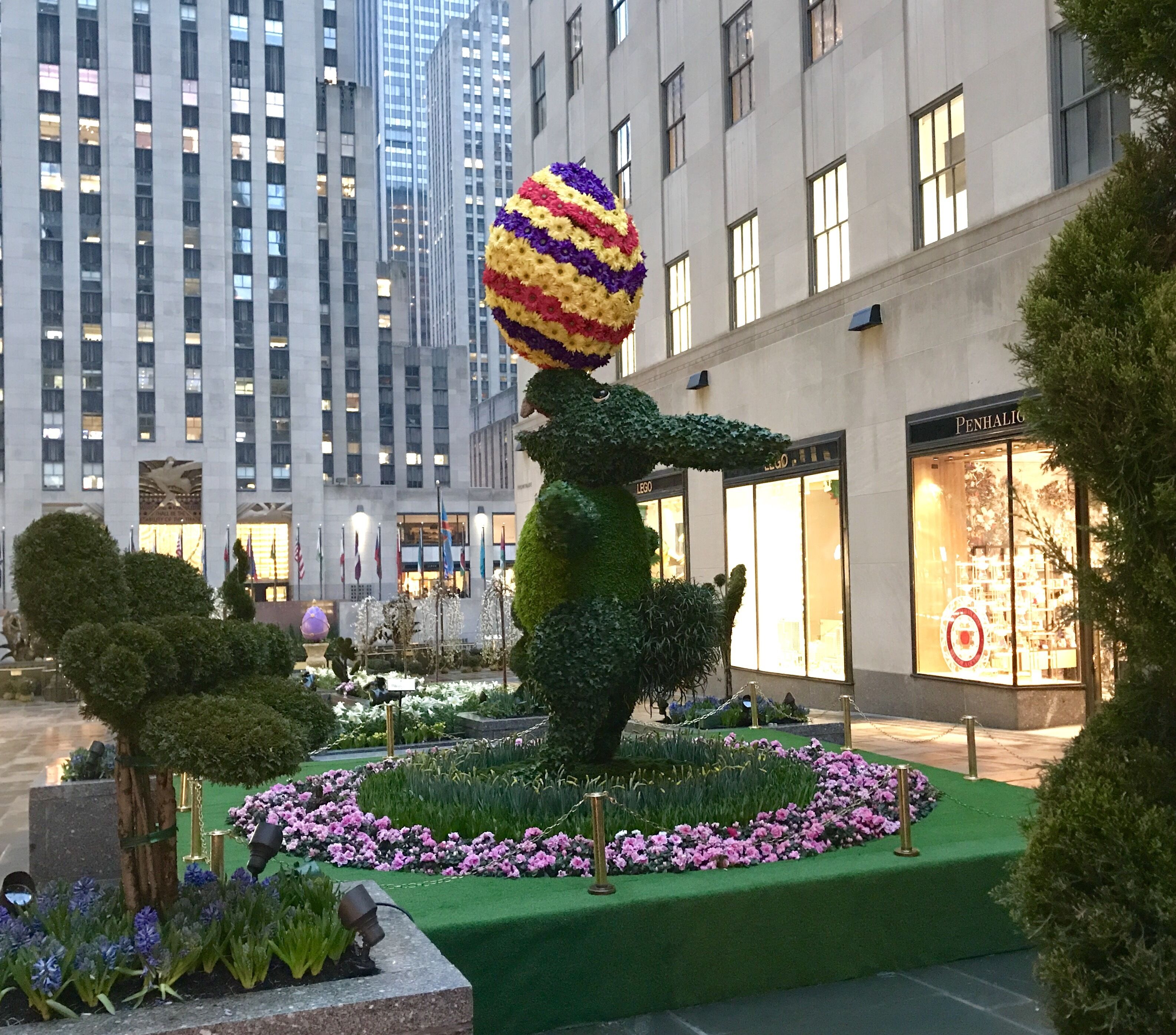 Um jardim bem cuidado com uma escultura floral esférica, rodeado por arbustos e flores aparados, em frente a um edifício com vitrine.