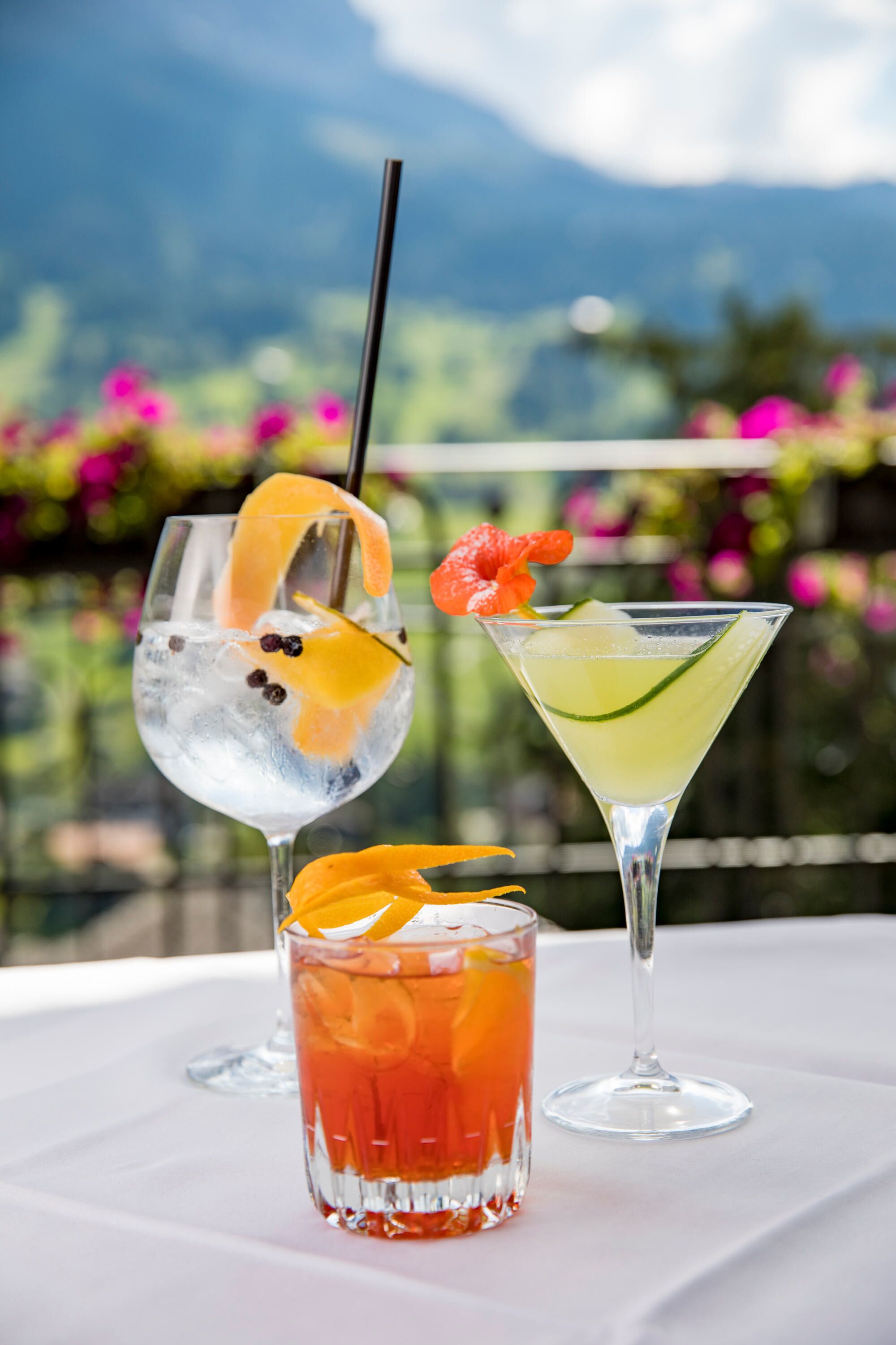 Dois cocktails sobre uma mesa, com montanhas e flores ao fundo.
