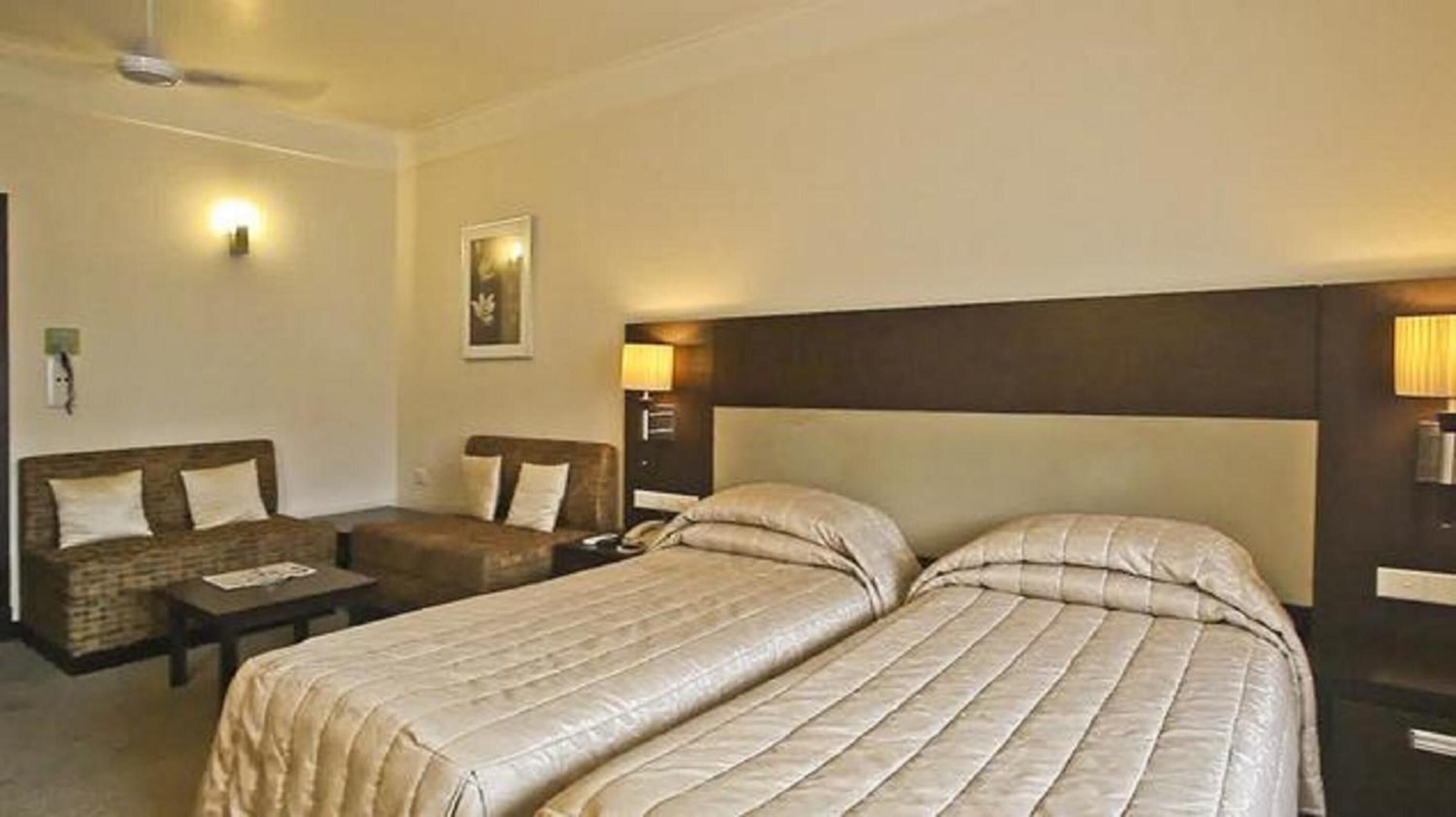 Quarto de hotel com duas camas, um sofá, uma mesinha e uma luminária.