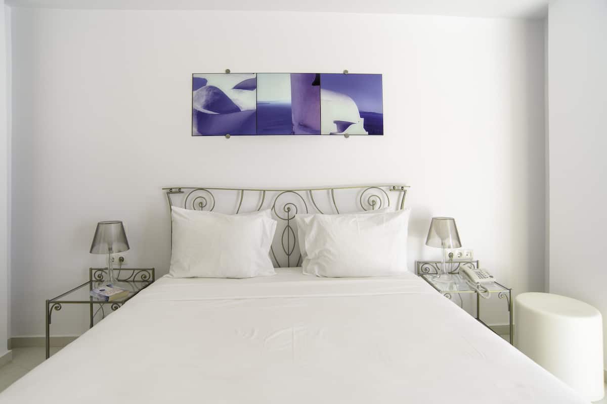Quarto de hotel bem arrumado com uma cama grande, duas mesas de cabeceira com abajures, uma mesinha com telefone e uma obra de arte na parede.