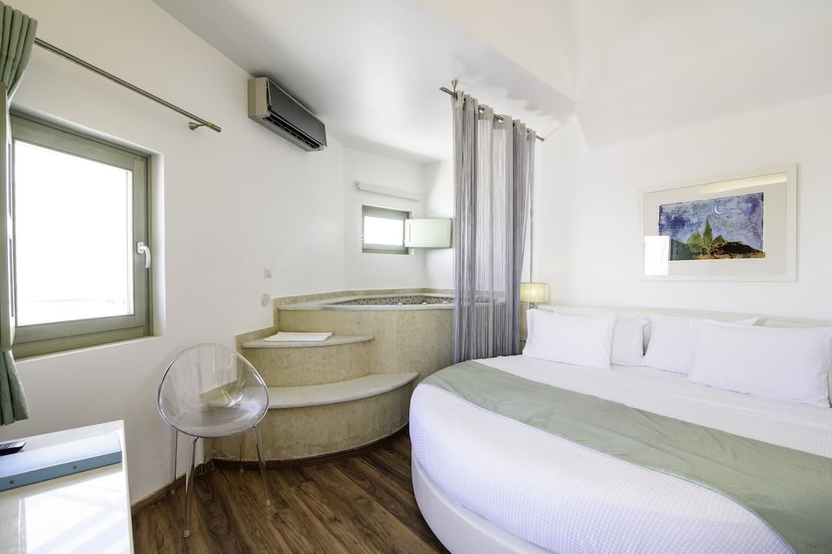 Quarto de hotel com cama, uma cadeira, uma mesinha, janela com cortinas, um quadro na parede e banheiro com pia e espelho.