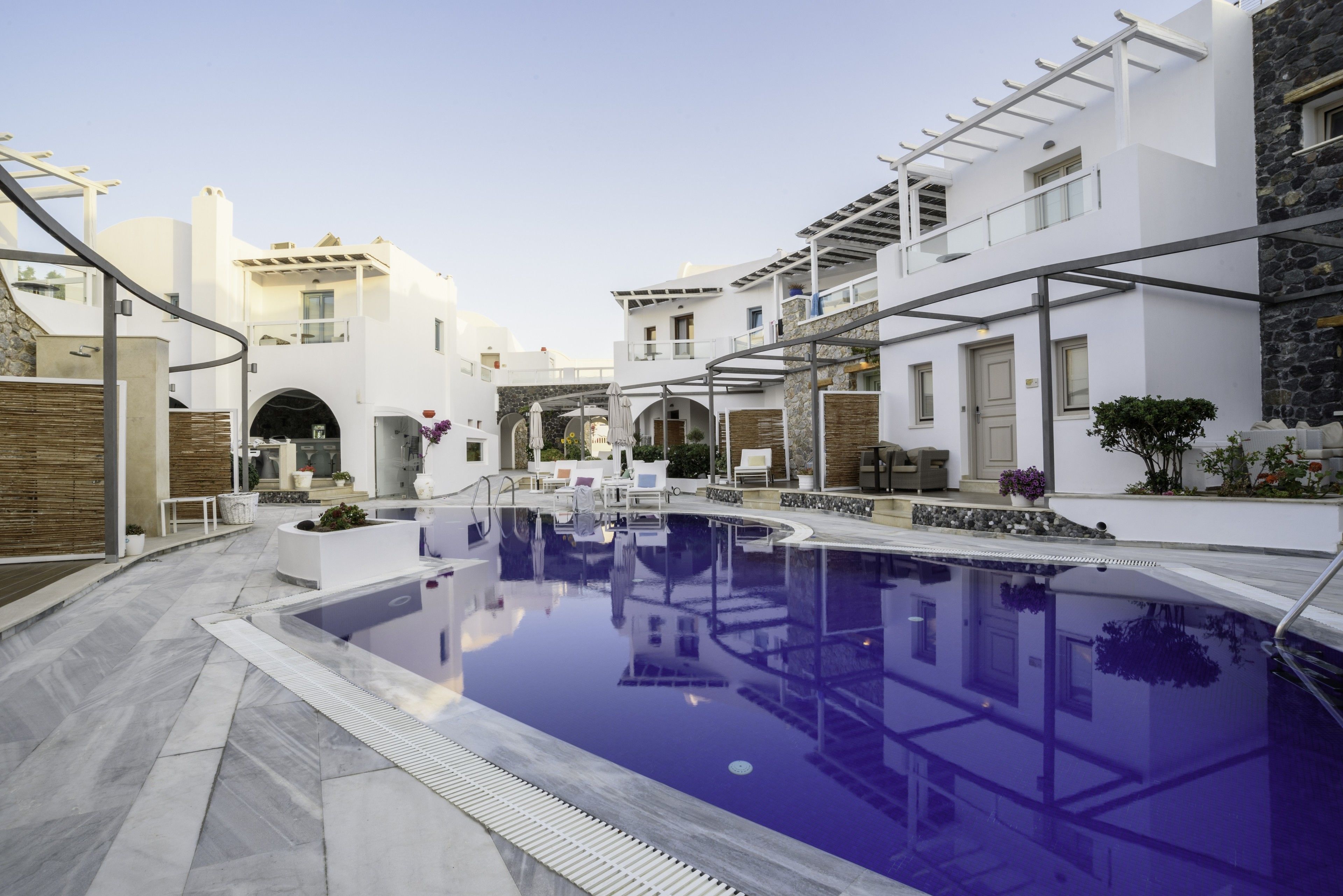 Uma área de piscina moderna com piscina transparente, edifícios brancos e mobiliário de exterior.