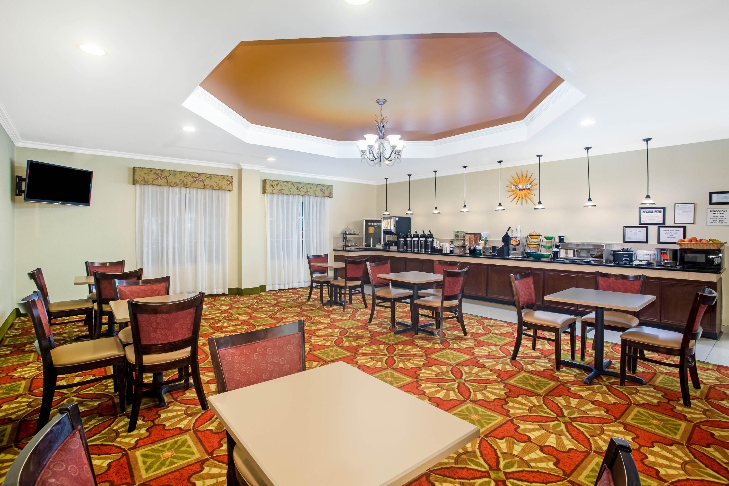 Uma área de refeições de hotel com bar, várias mesas e cadeiras, e um tapete estampado.