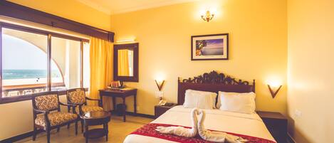 Quarto de hotel com cama, duas cadeiras, uma mesinha, mesa de cabeceira, ventoinha no teto, janela com cortinas e um quadro na parede.
