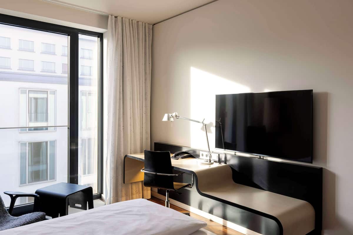Quarto de hotel moderno com cama, escrivaninha com cadeira, televisão de tela plana e uma grande janela com vista para a cidade.