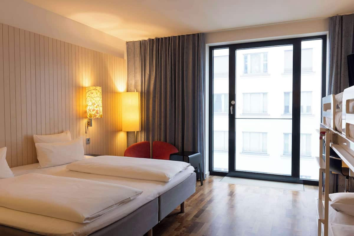 Quarto de hotel com uma cama grande, uma escrivaninha, uma cadeira, um abajur e vista da cidade através de uma janela ampla.
