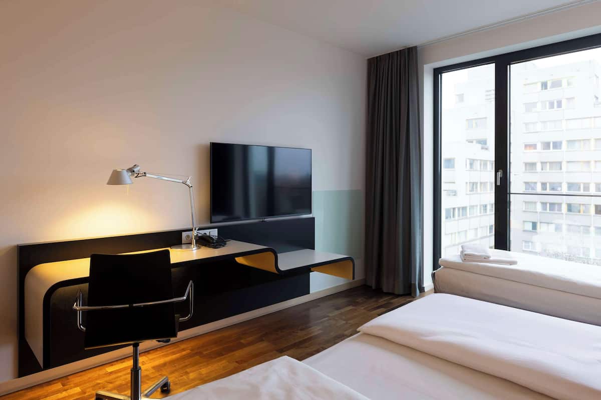 Quarto de hotel com cama, escrivaninha com cadeira, televisão e janela com vista para prédios.