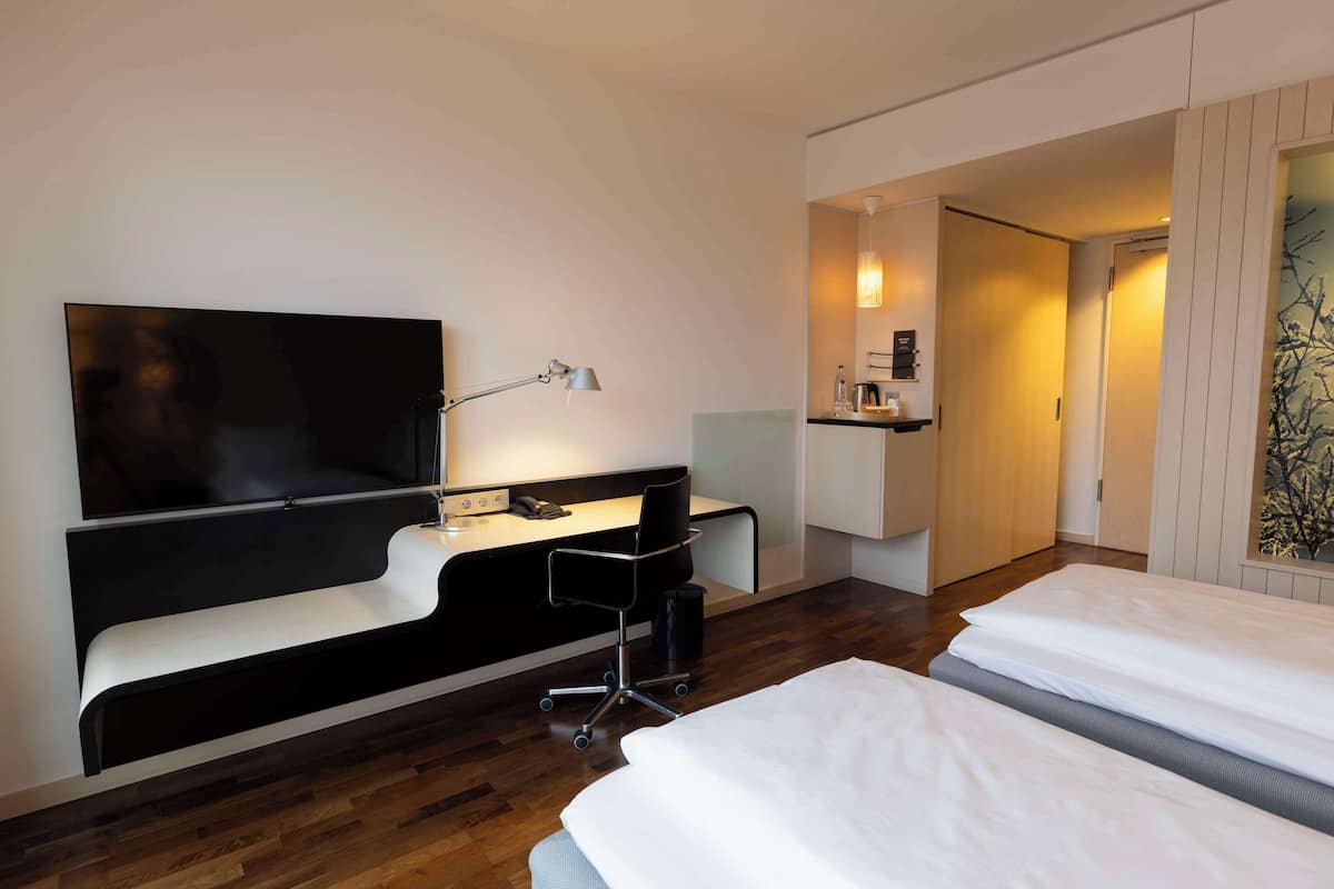 Quarto de hotel moderno com televisão de tela plana, escrivaninha com cadeira, cama com duas almofadas e um pequeno espaço de trabalho com luminária.