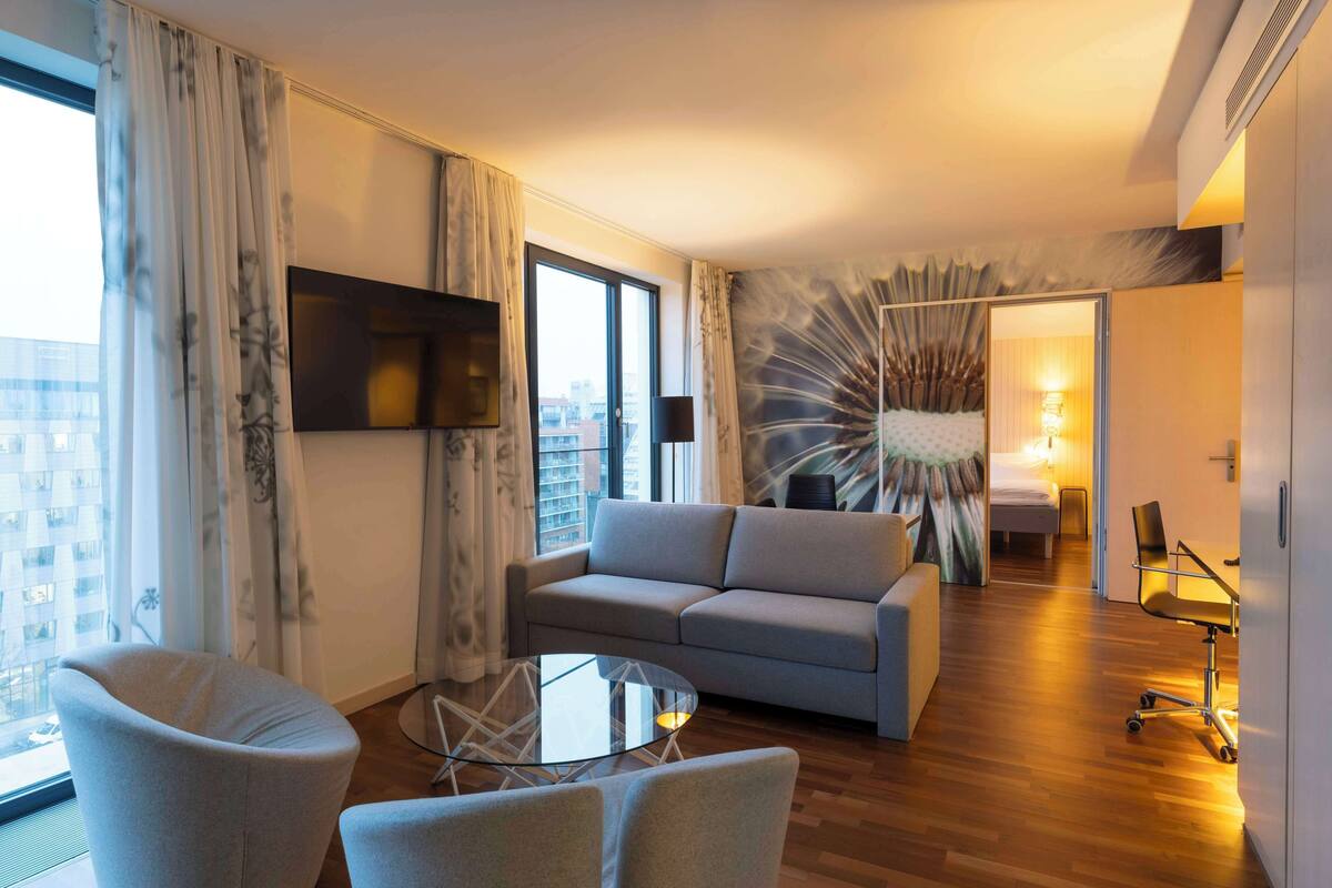 Quarto de hotel moderno com um sofá cinzento, uma mesa de centro de vidro e uma janela ampla com cortinas.