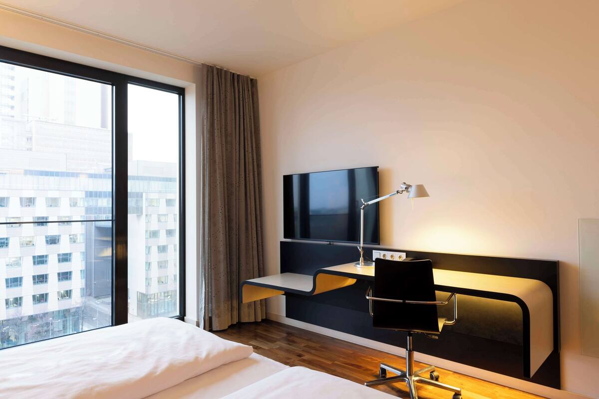 Quarto de hotel com uma grande janela, uma cama, uma escrivaninha com cadeira, uma televisão e um abajur.