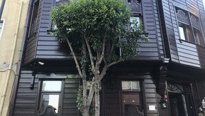 Um edifício de vários andares com características arquitetónicas tradicionais, incluindo janelas decorativas e uma árvore em frente.