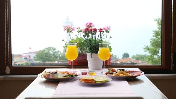 Uma mesa de café da manhã com pratos de comida, dois copos de suco de laranja e um vaso de planta perto de uma janela com vista para edifícios e uma bandeira.