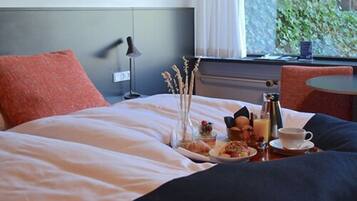 Quarto de hotel com cama, mesa de cabeceira com abajur, pequena mesa com itens de café da manhã e vista para áreas verdes lá fora.