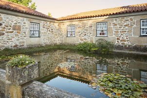 Um edifício de pedra com telhado de telhas, um lago com folhas de nenúfar e uma pequena ponte.