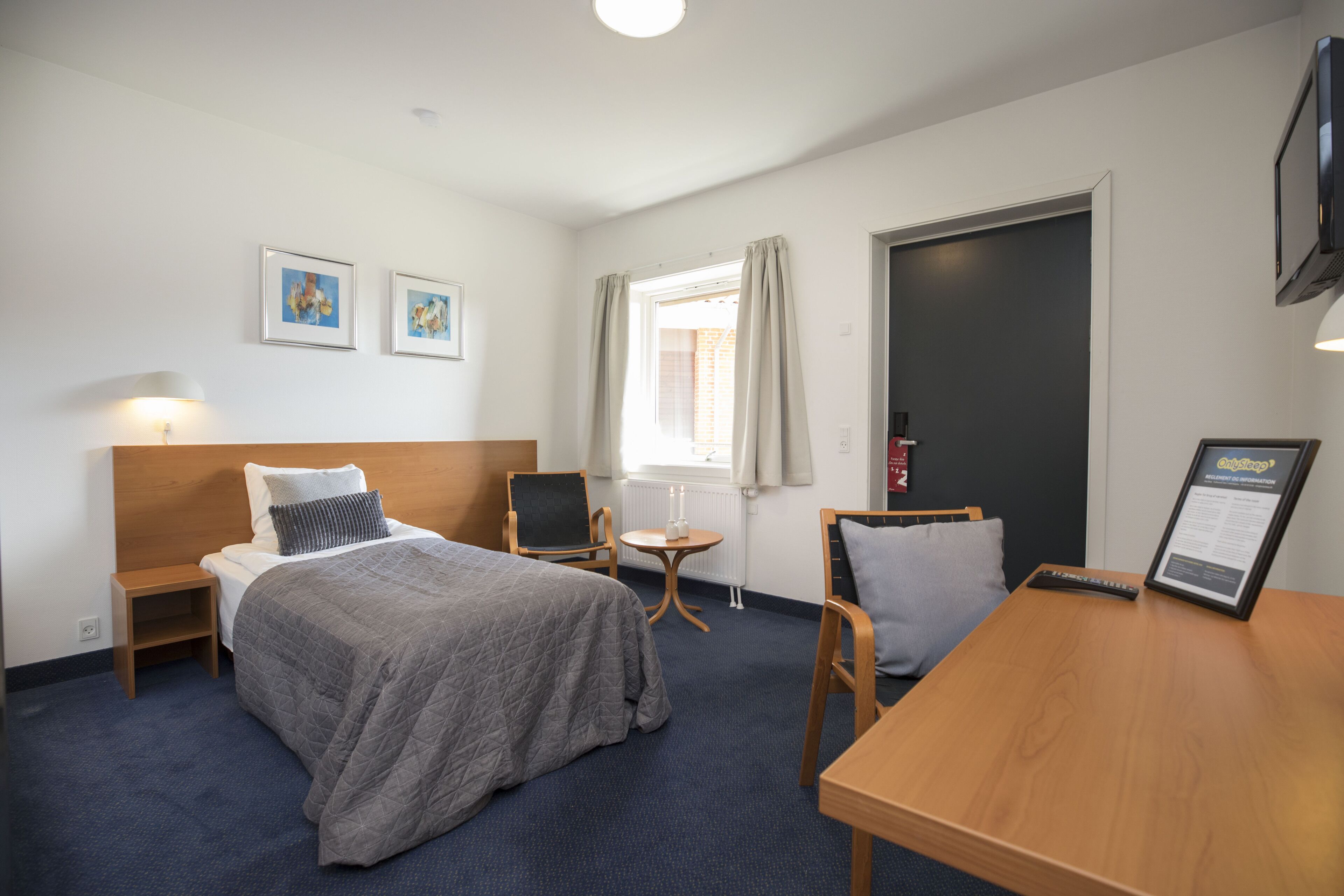 Quarto de hotel com uma cama, uma escrivaninha, uma cadeira, uma mesinha e uma televisão.