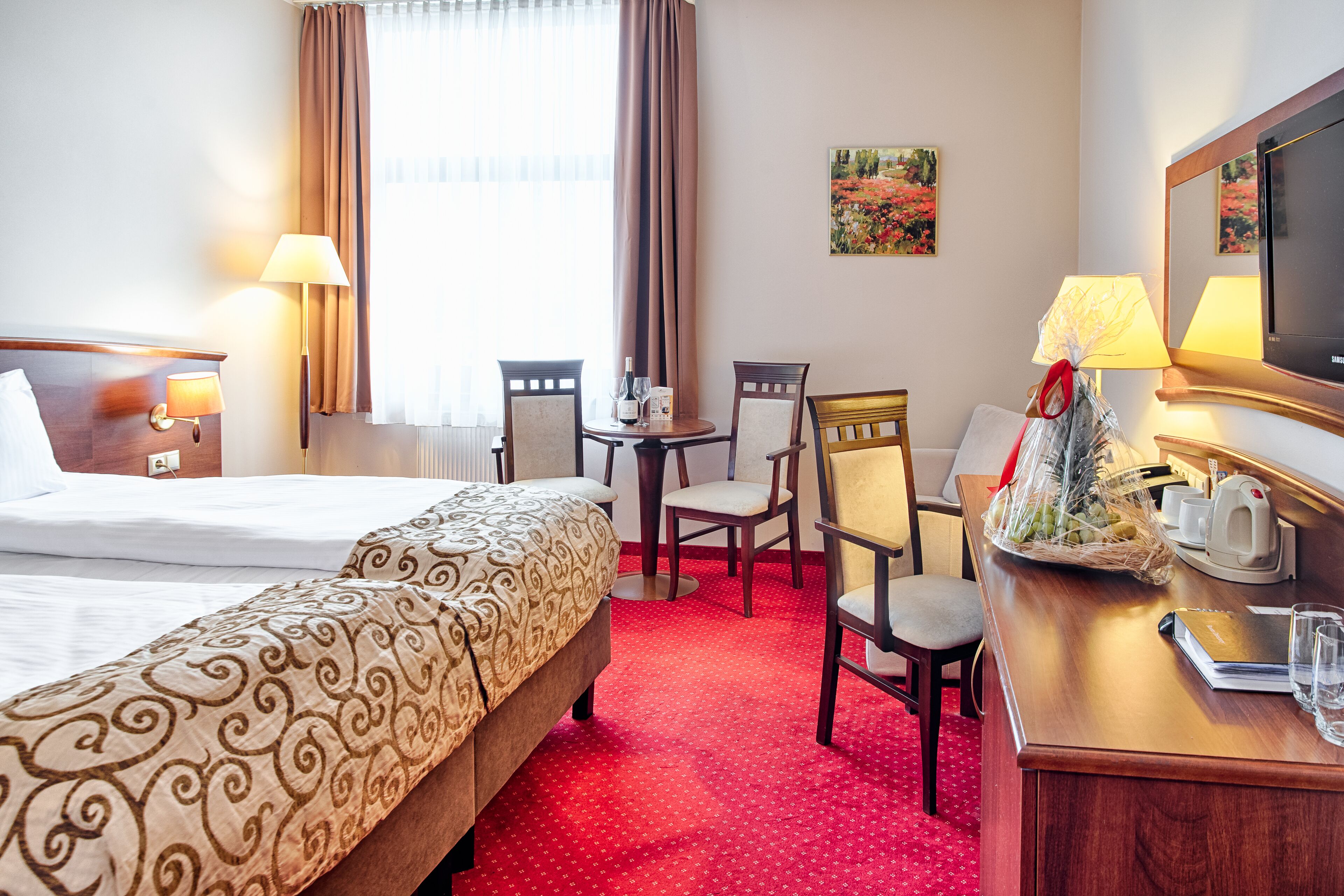 Quarto de hotel com uma cama, duas cadeiras, uma mesa pequena com um abajur, uma televisão e um tapete vermelho.