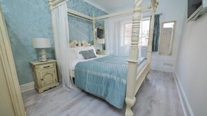 Uma cama de dossel com um cobre-leito azul, uma mesa de cabeceira com abajur e um espelho na parede.