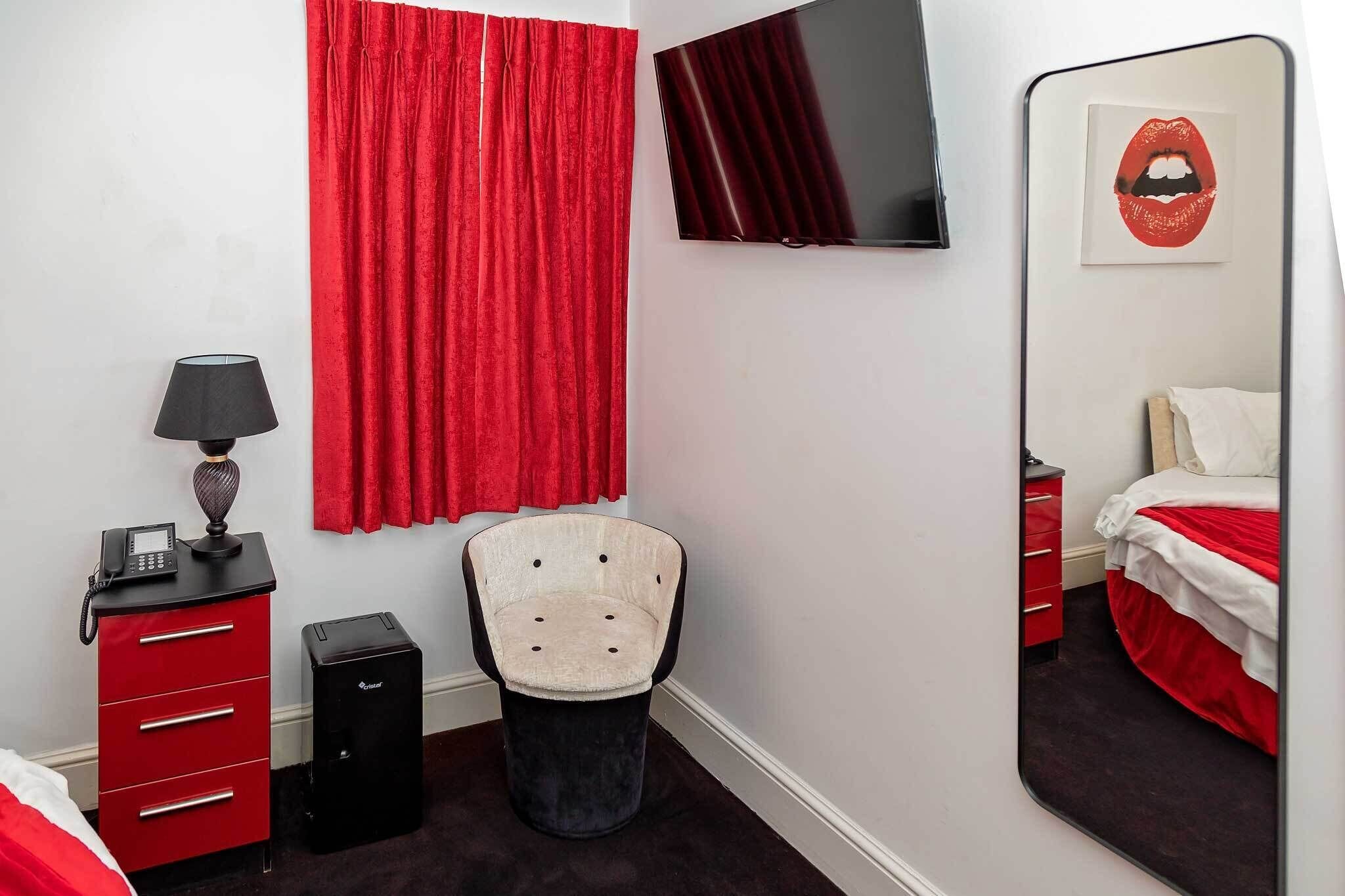 Quarto de hotel com cama vermelha, secretária preta com luminária, cadeira, pequeno alto-falante preto e TV fixada na parede.