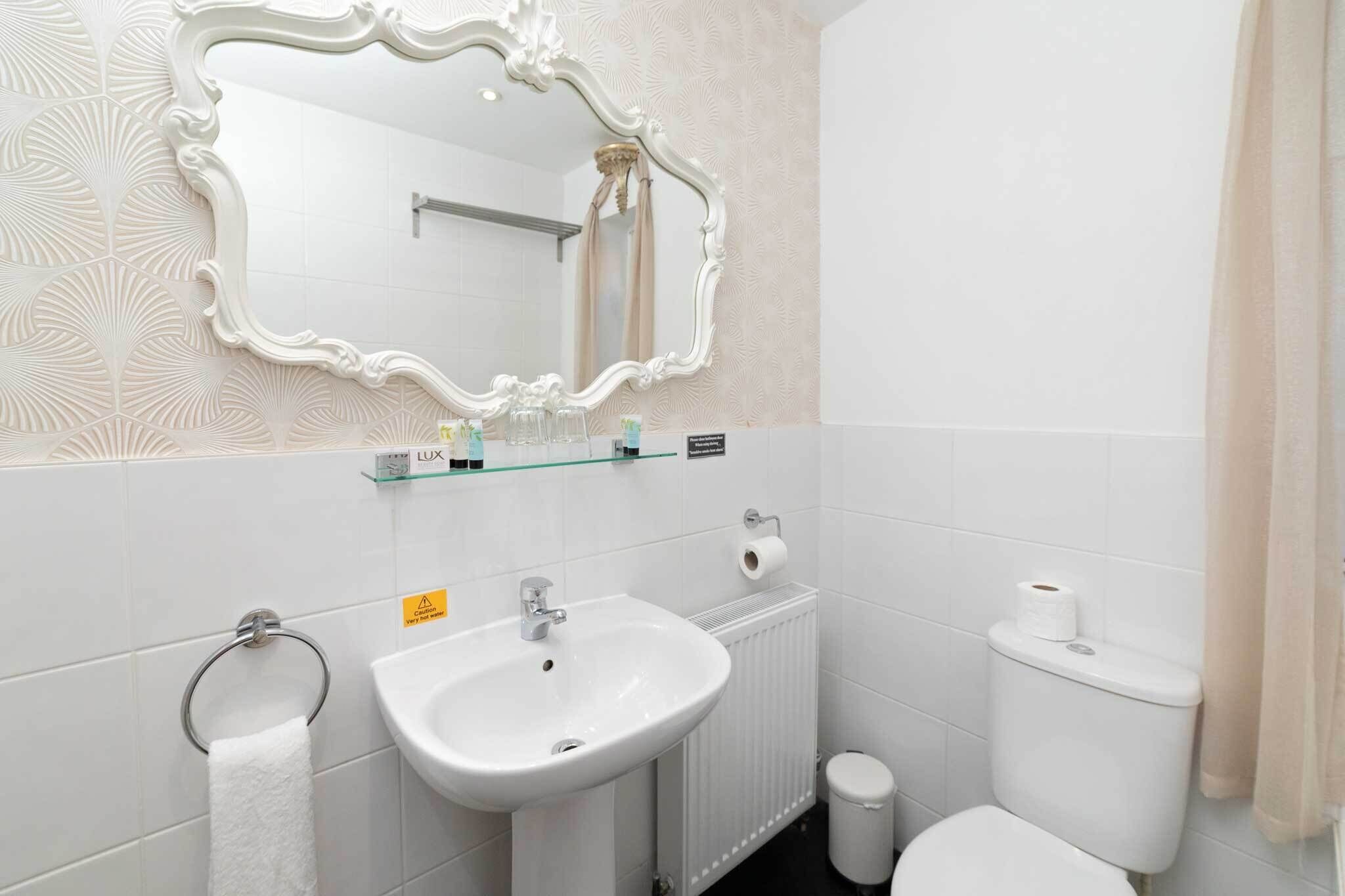 Casa de banho com lavatório branco, espelho com moldura ornamentada, sanita e porta-toalhas.