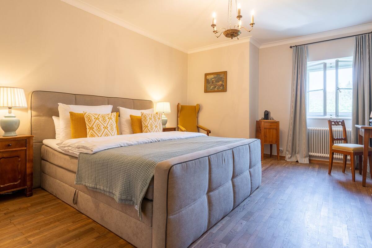 Quarto com uma cama grande, mesas de cabeceira de madeira, uma cadeira, escrivaninha e janela com cortinas.